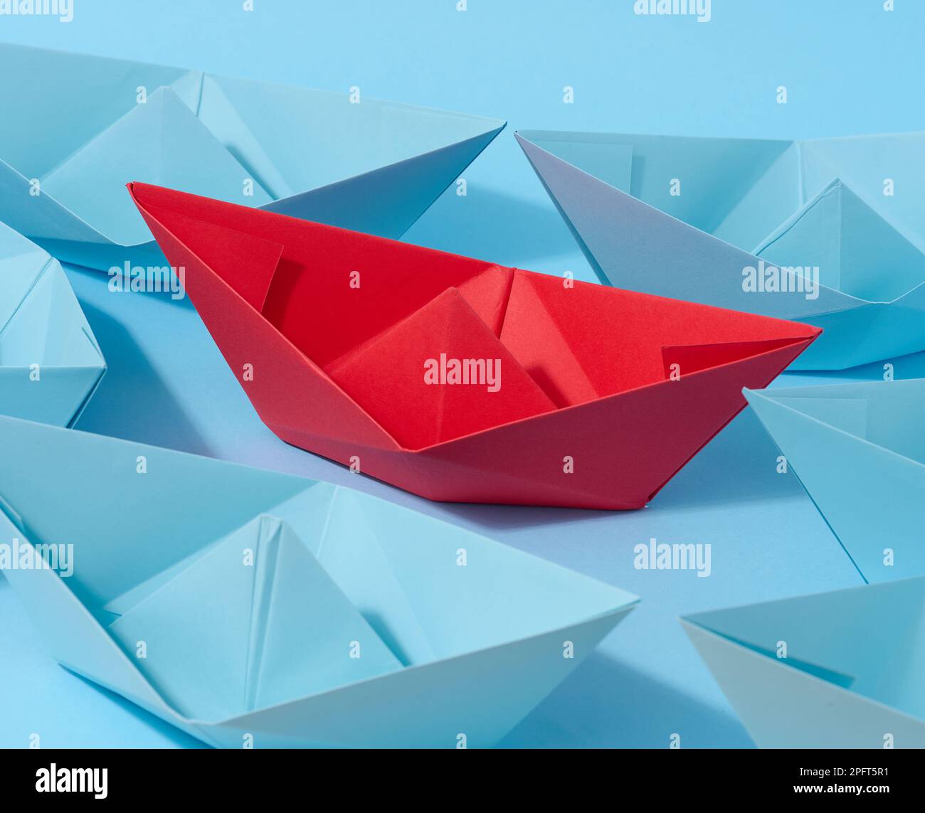 A red paper boat stands in front of a group of blue paper boats, a ...