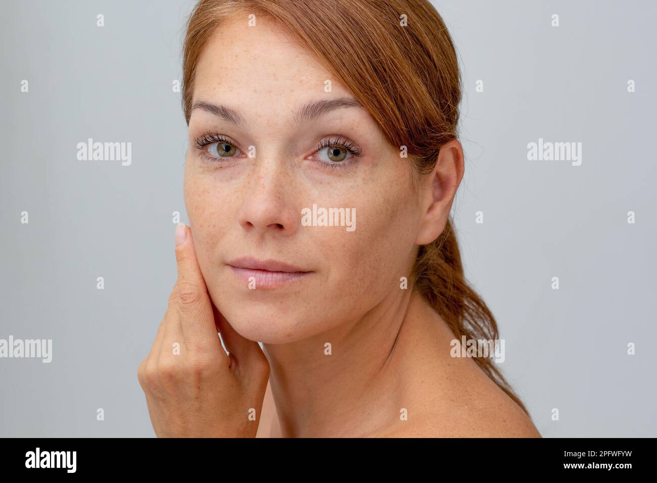 Portrait of middle aged woman with reddish hair holding hand on cheek looking at camera on gray ...