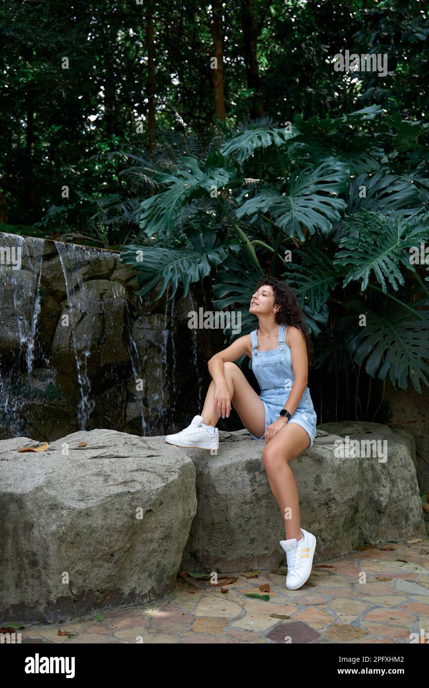 A beautiful girl poses on rocks near water in the jungle among plants ...