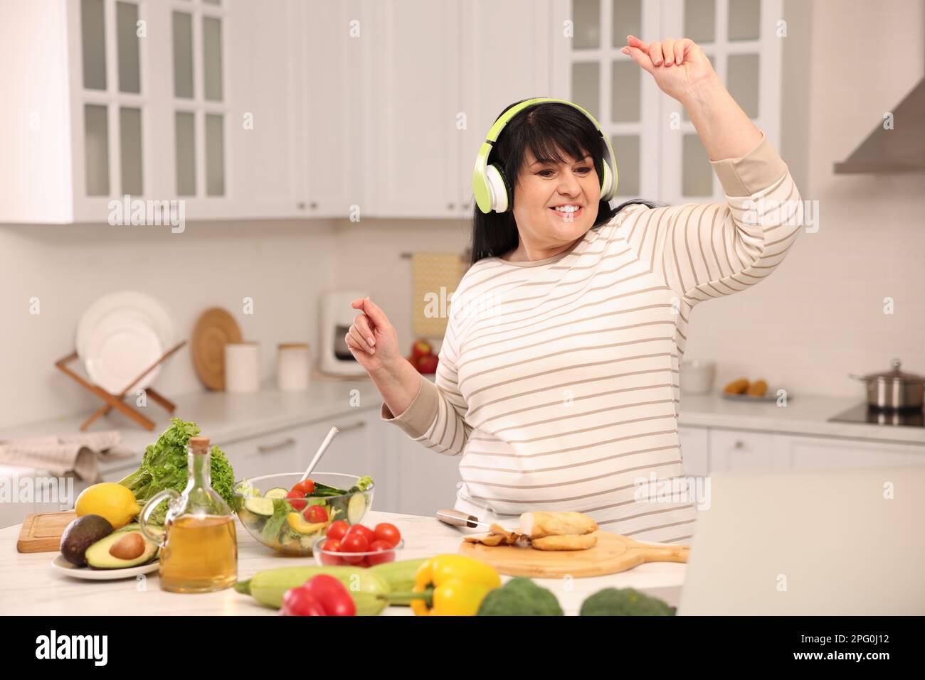Happy overweight woman with headphones dancing while cooking in kitchen ...
