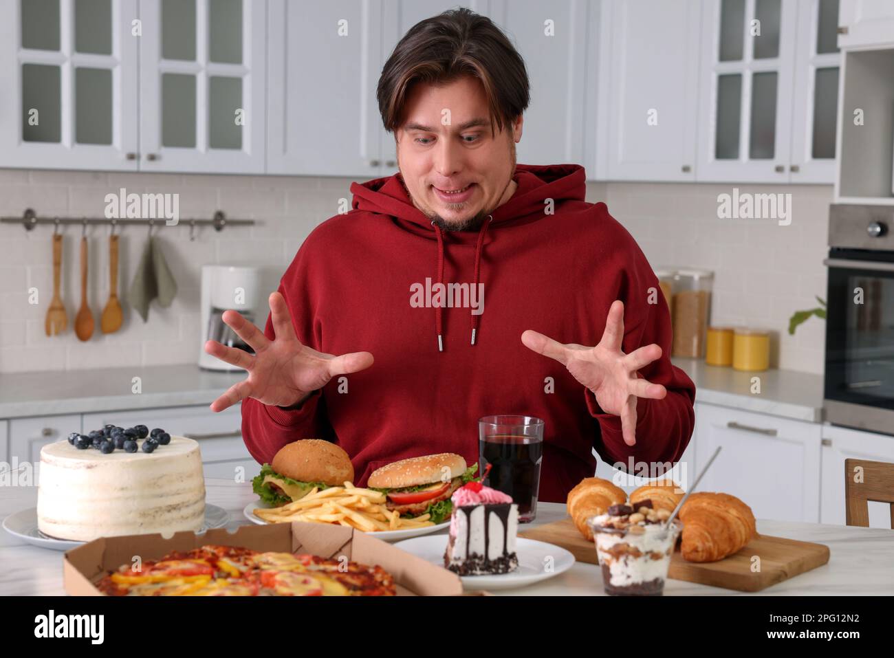 Hungry overweight man at table with sweets and fast food in kitchen ...