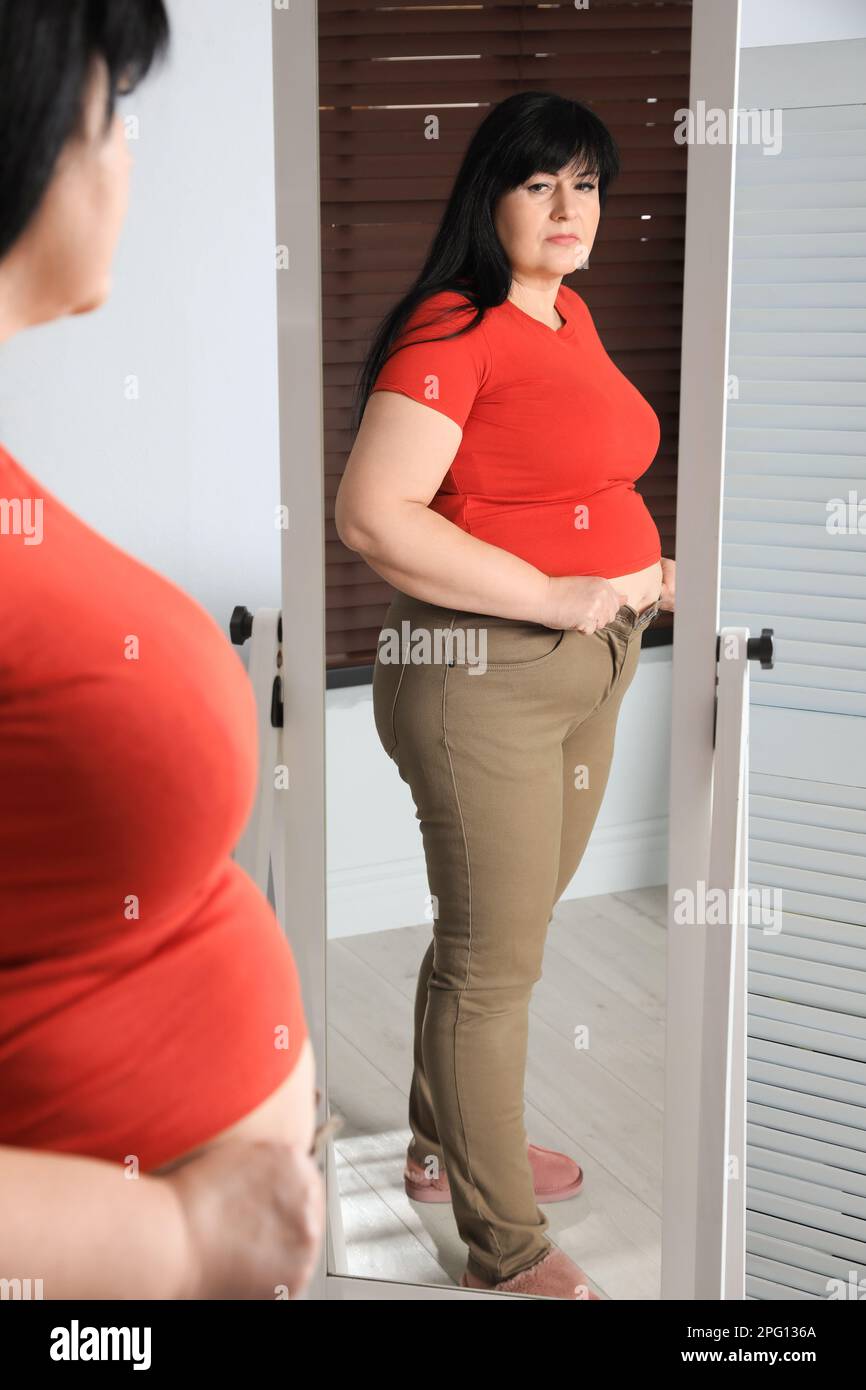 Overweight woman trying to button up tight trousers in front of mirror ...