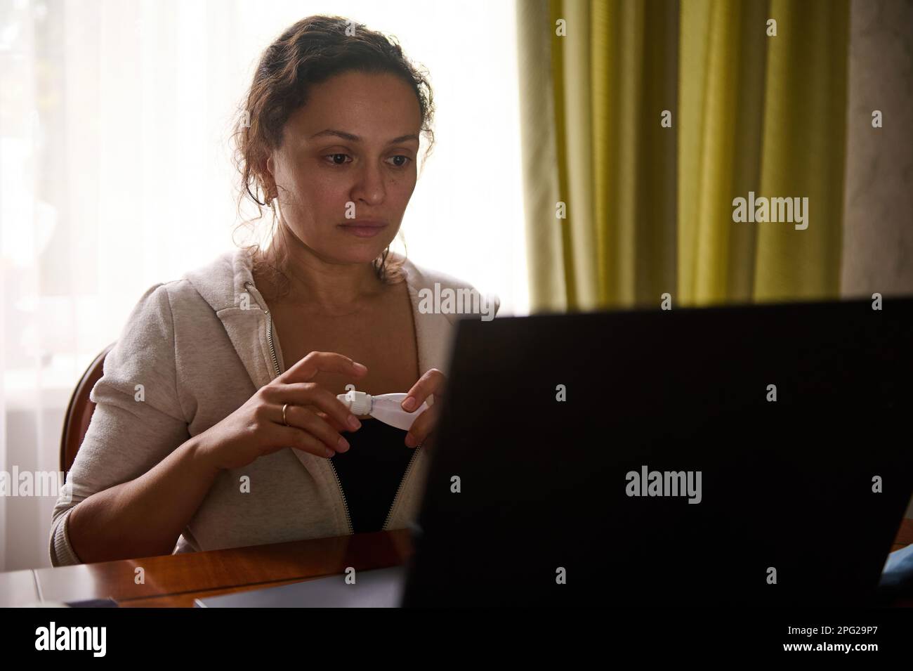 Female entrepreneur sitting at laptop and holding medicated eye drops ...
