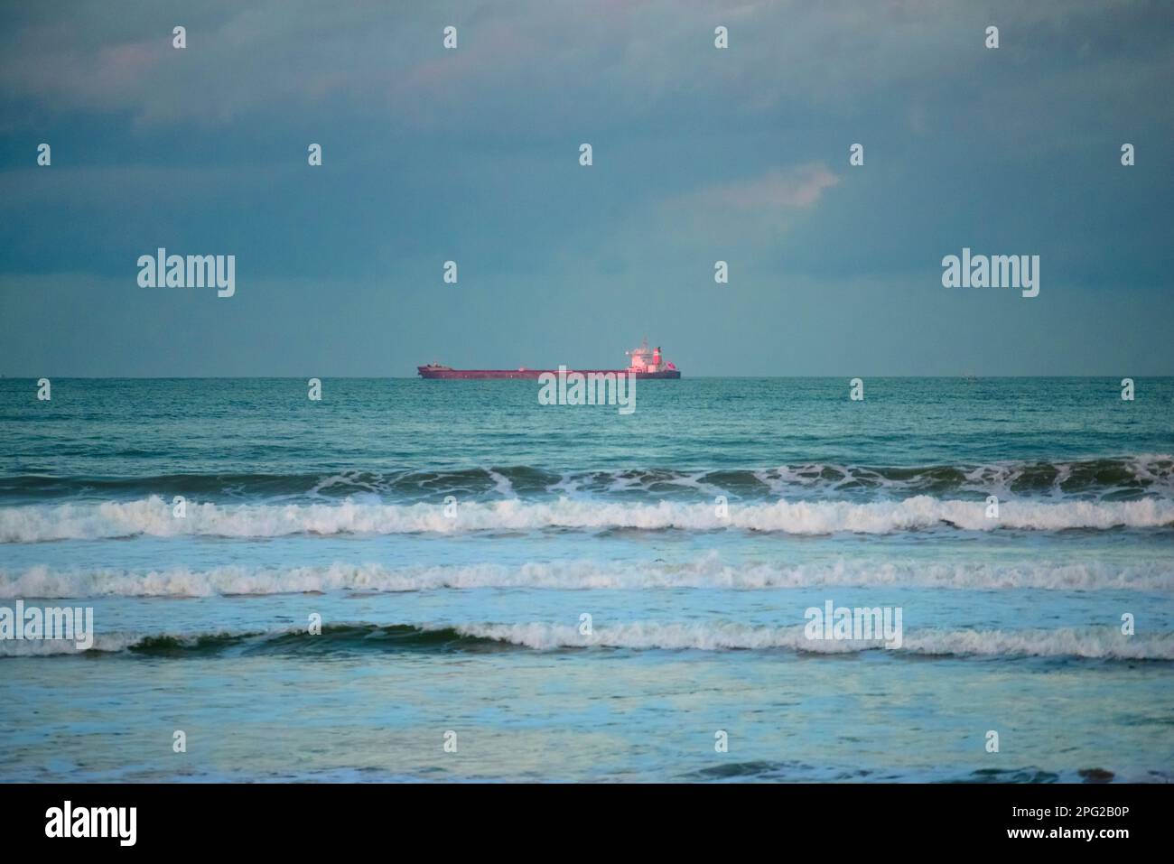 A cargo ship over the horizon under grey blue sky and sea with waves ...