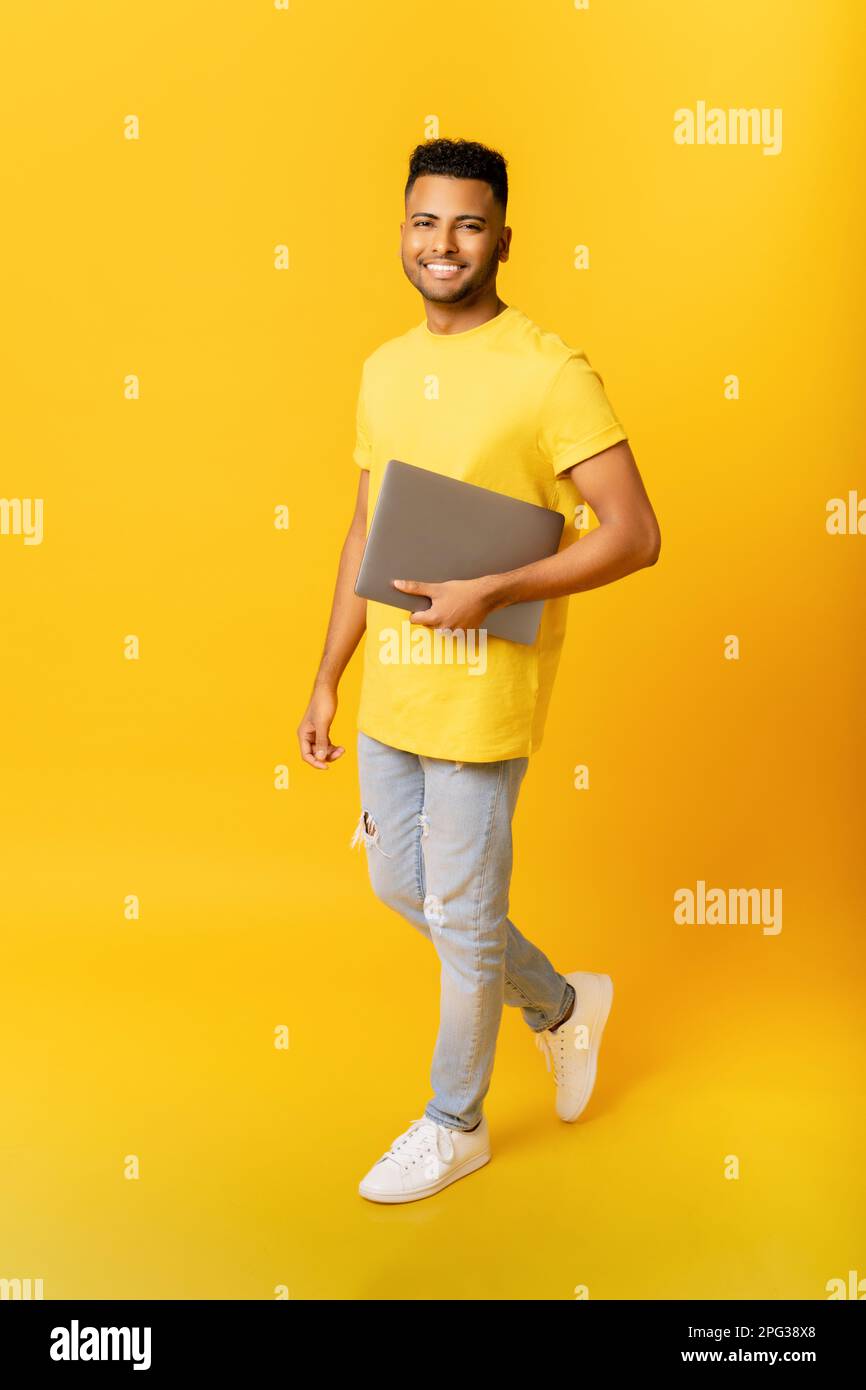 Cheerful Indian man carrying laptop computer isolated on yellow. Happy ...