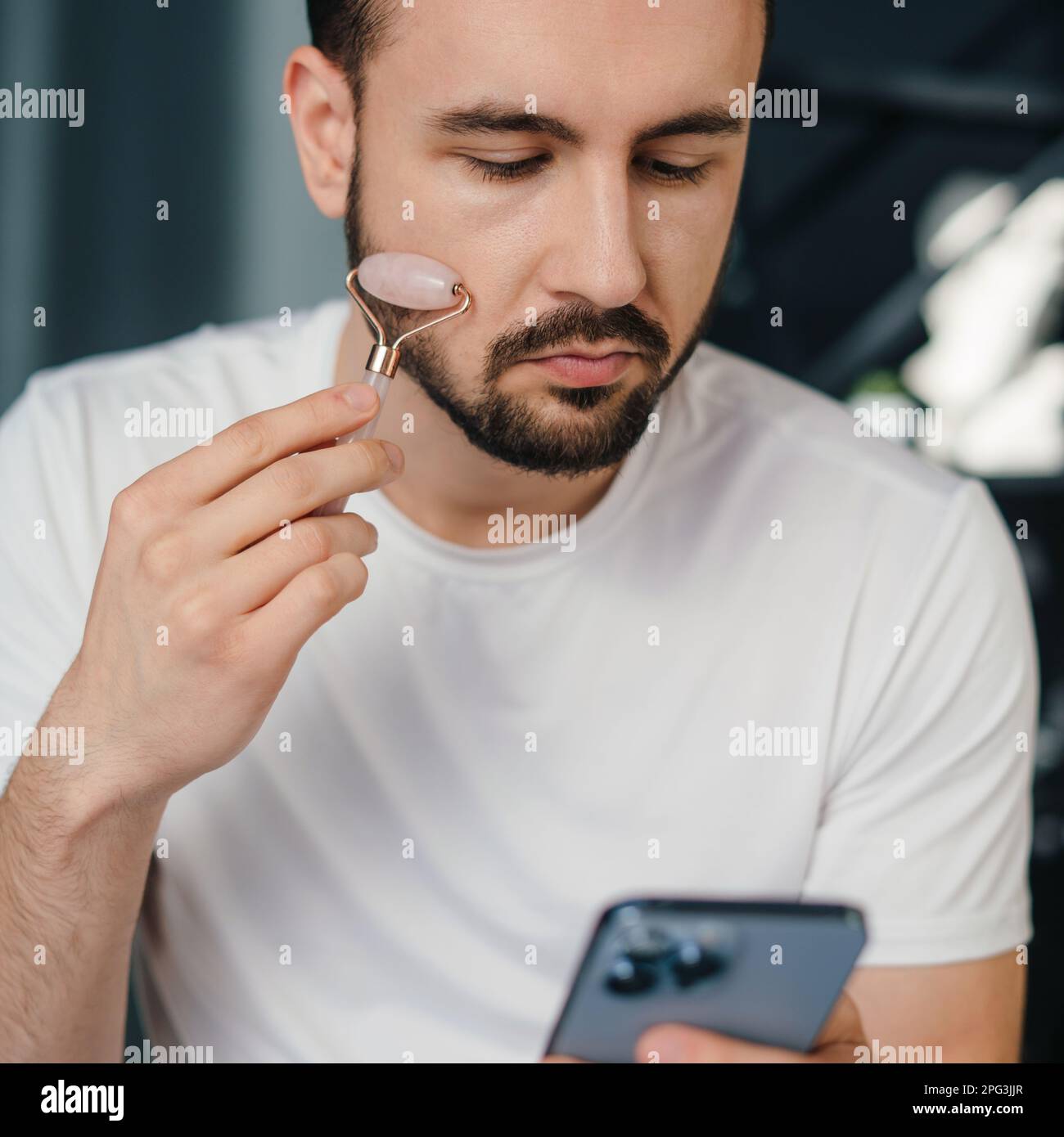 Calm man standing at home using roller massager on his face, looking at ...