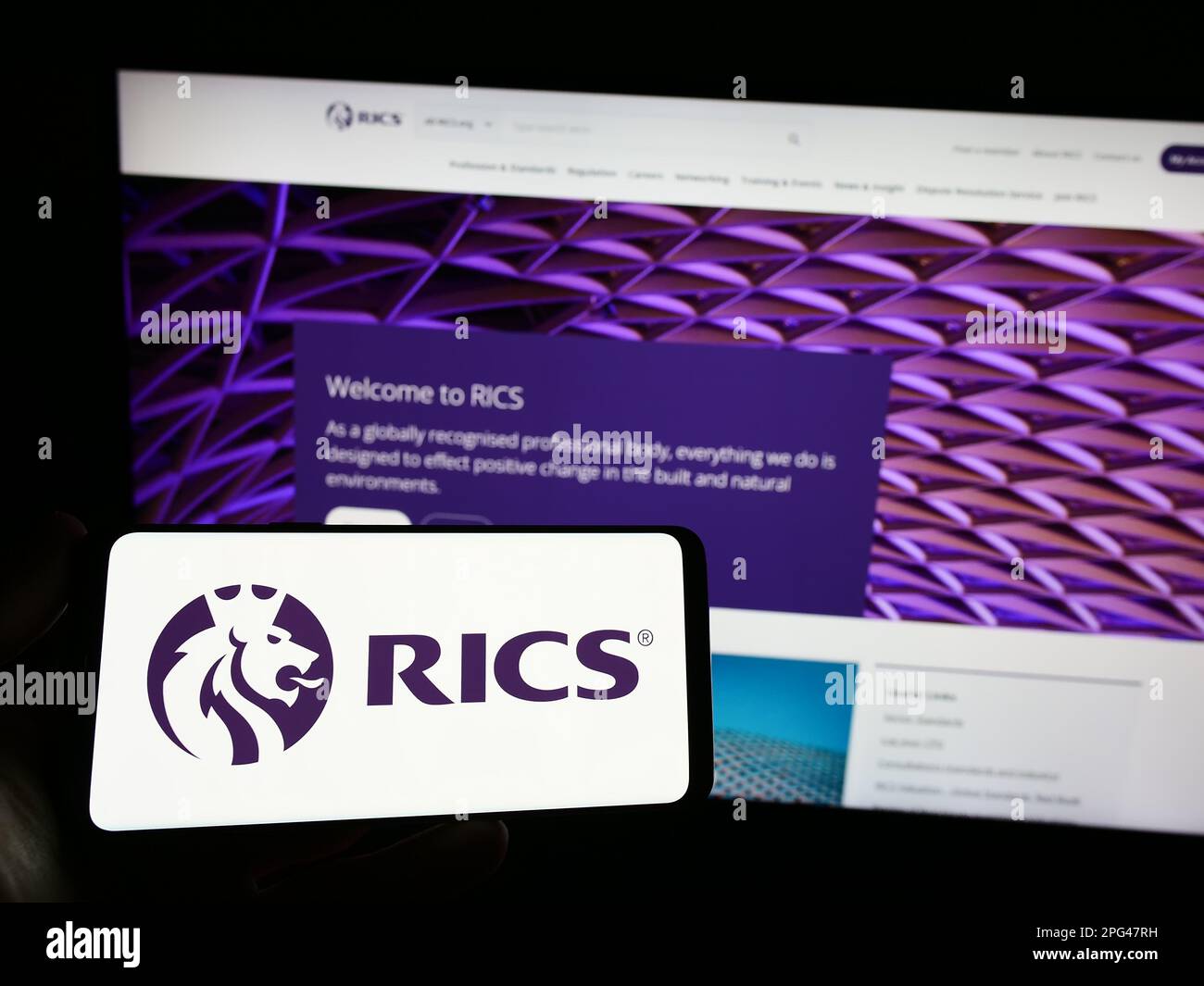 Person holding smartphone with logo of Royal Institution of Chartered Surveyors (RICS) on screen ...