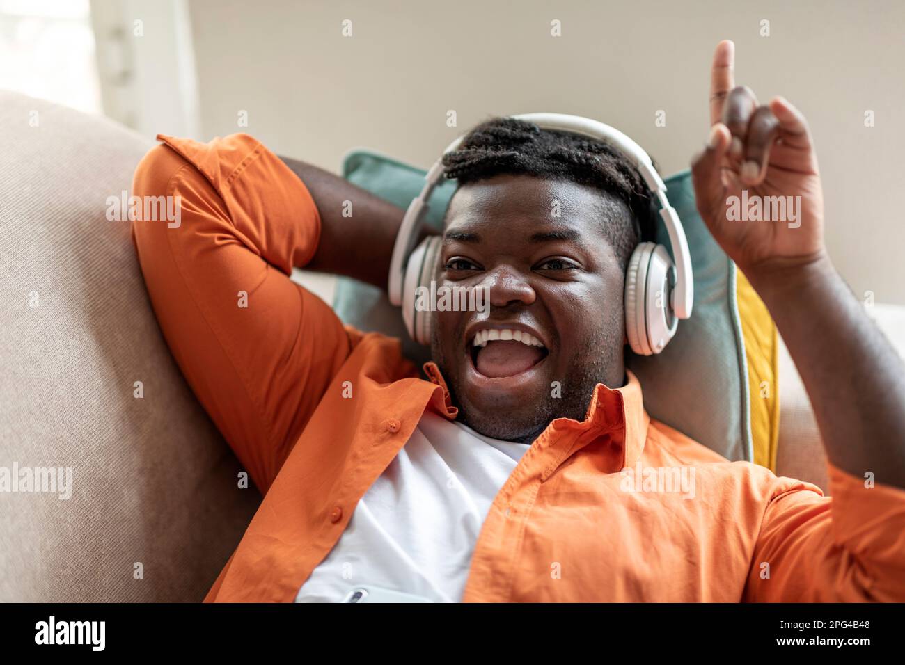 Joyful black guy reclining on sofa, singing songs, using headphones ...