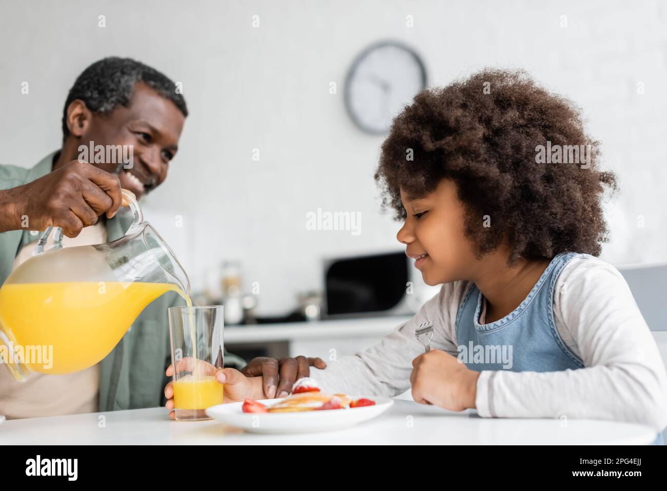 happy african american girl looking at glass while grandfather pouring orange juice during ...