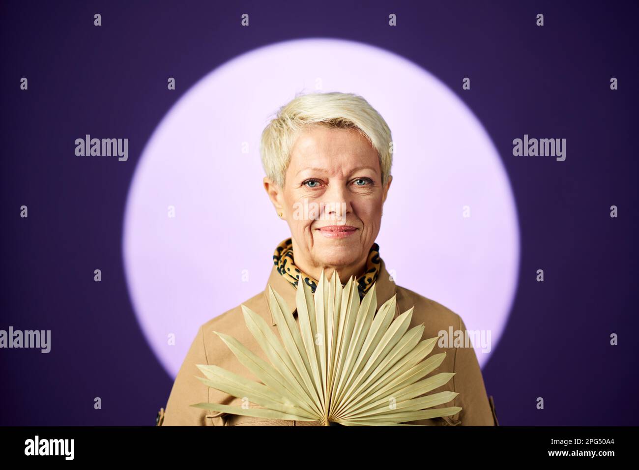 Mature blond woman with beige fan by her chest standing against large ...