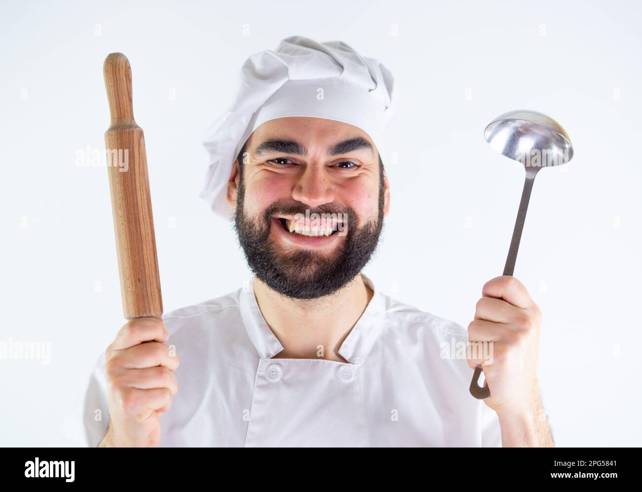 Young male chef showing a metal ladle and a wooden rolling pin while ...
