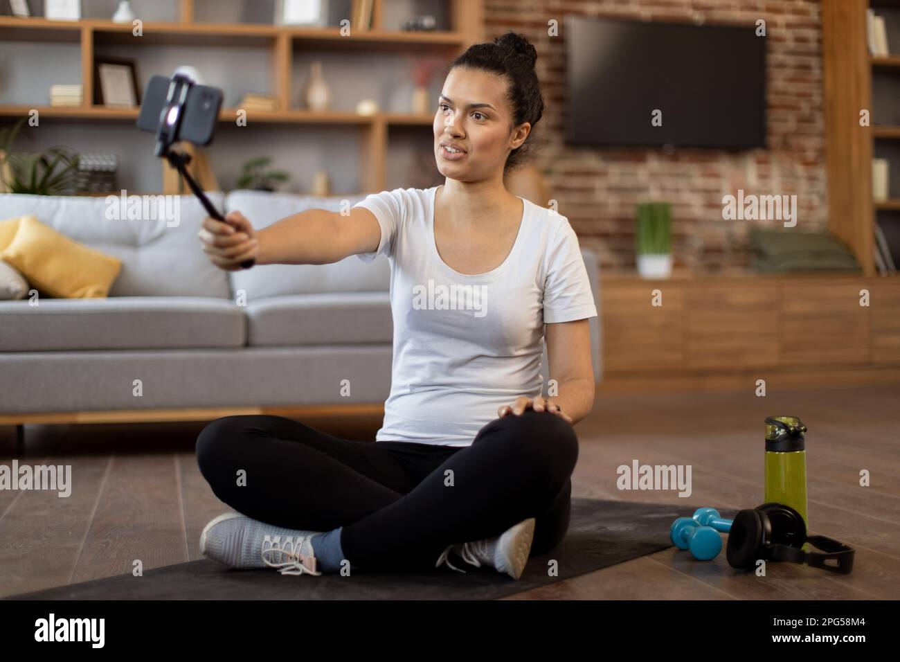 Young active multiracial woman in sportswear using cell phone on tripod ...