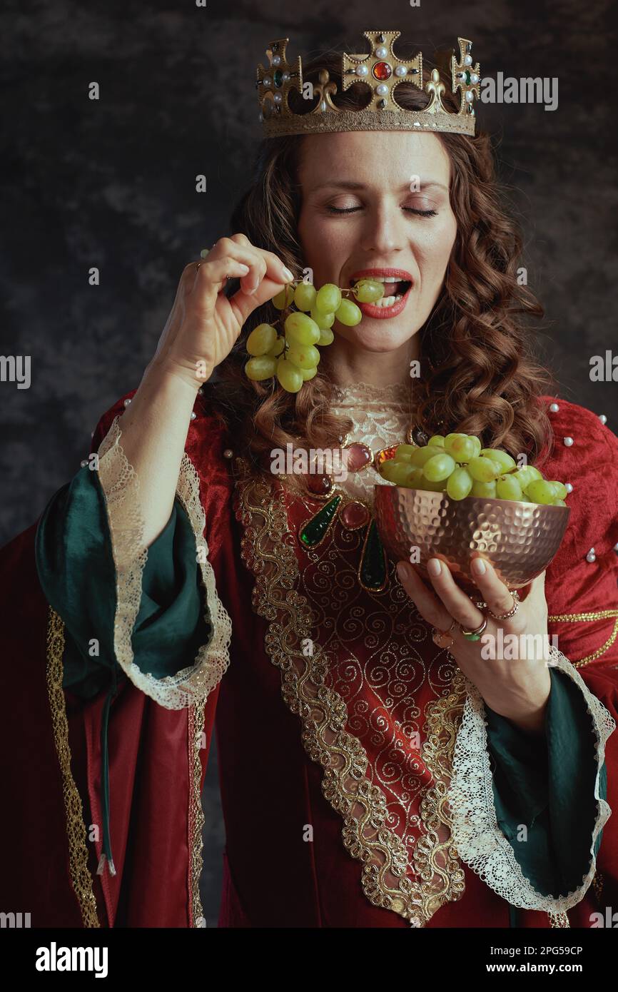 medieval queen in red dress with plate of grapes and crown on dark gray ...