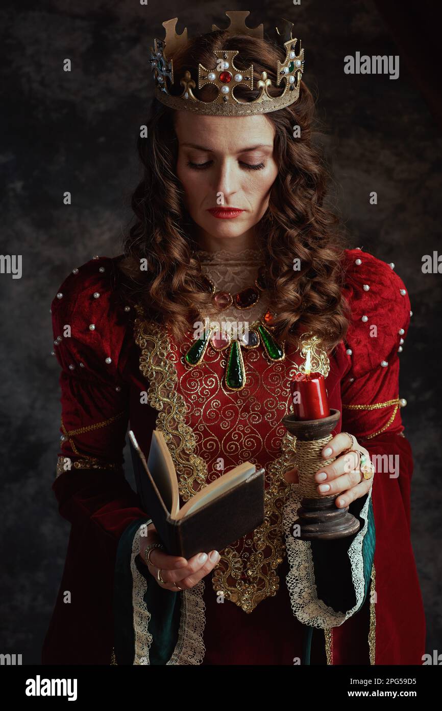 medieval queen in red dress with book, candle and crown on dark gray ...