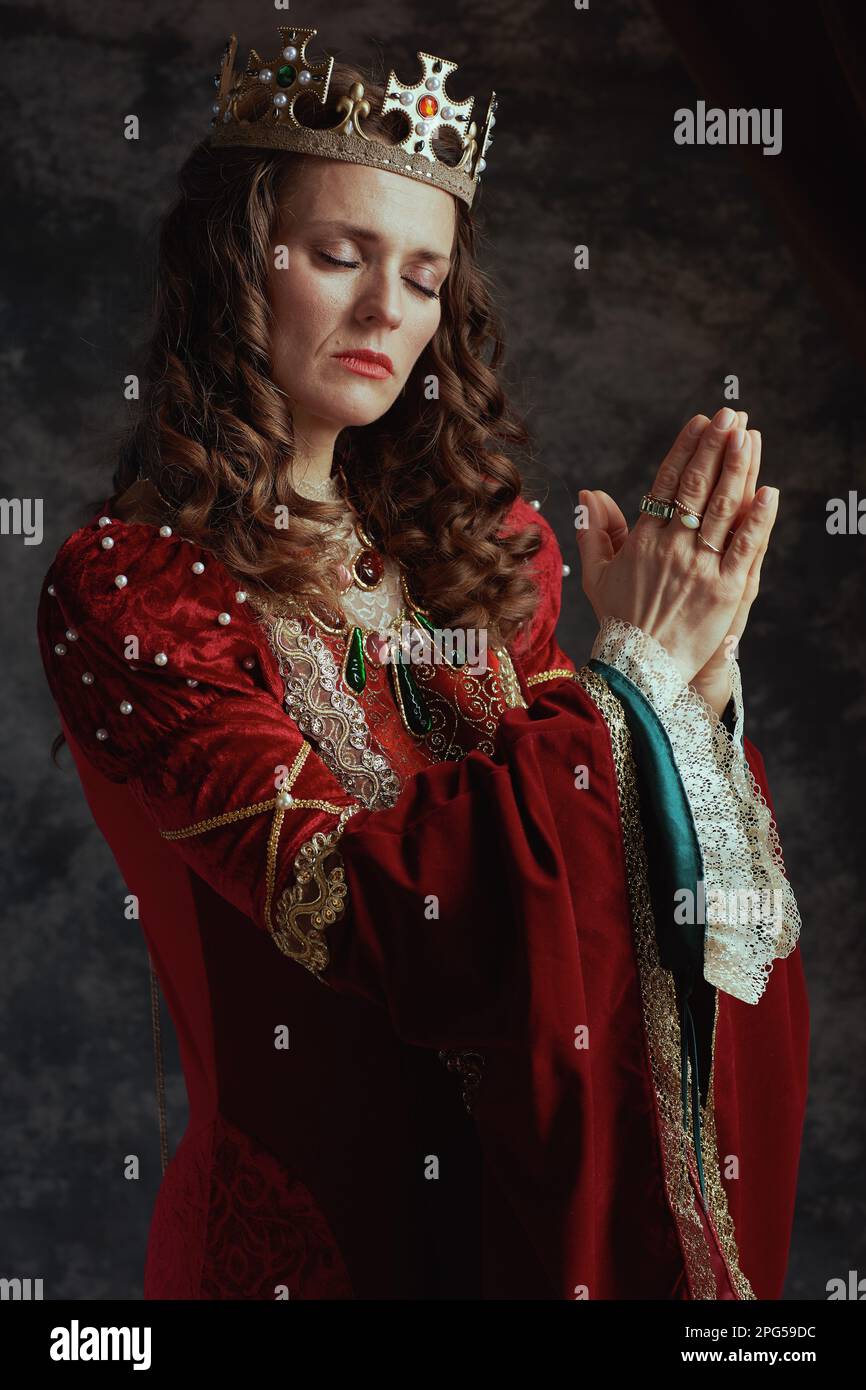 medieval queen in red dress with crown praying on dark gray background ...