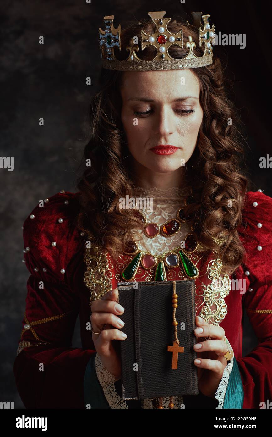 medieval queen in red dress with book, rosary and crown on dark gray ...