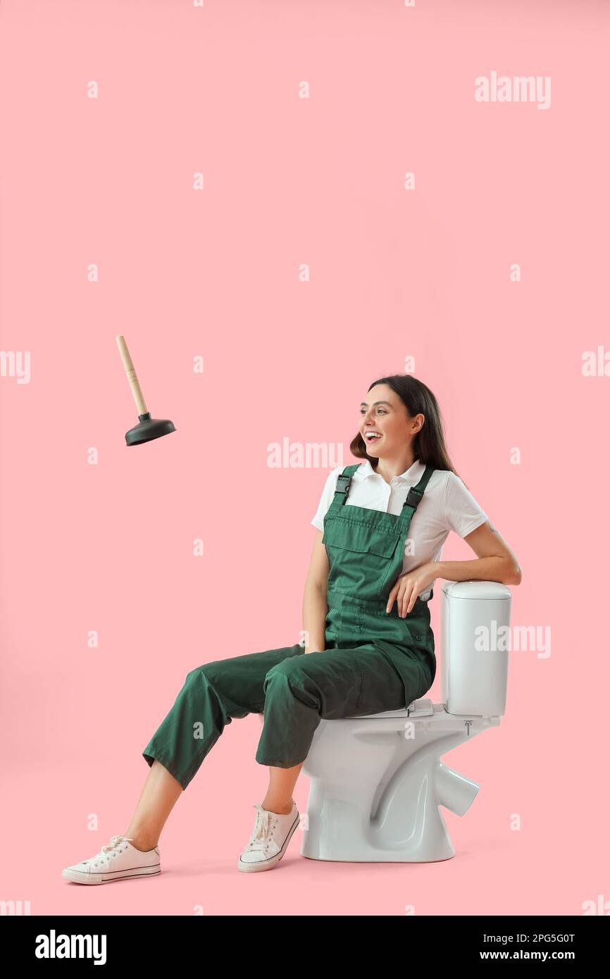 Female plumber sitting on toilet bowl and flying plunger against pink ...