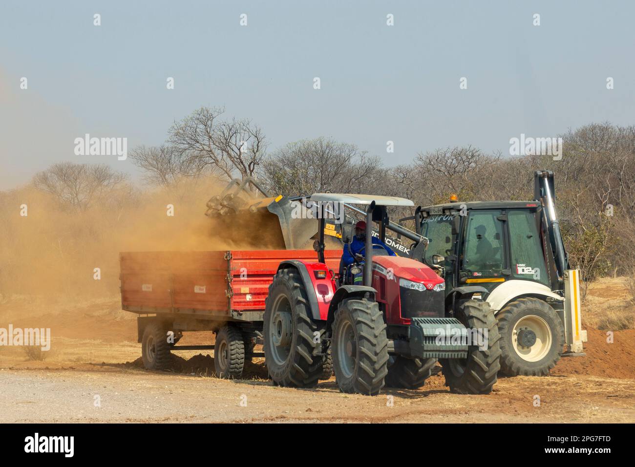 A back actor filling red soil into a tractor-drawn trailer bin on a ...