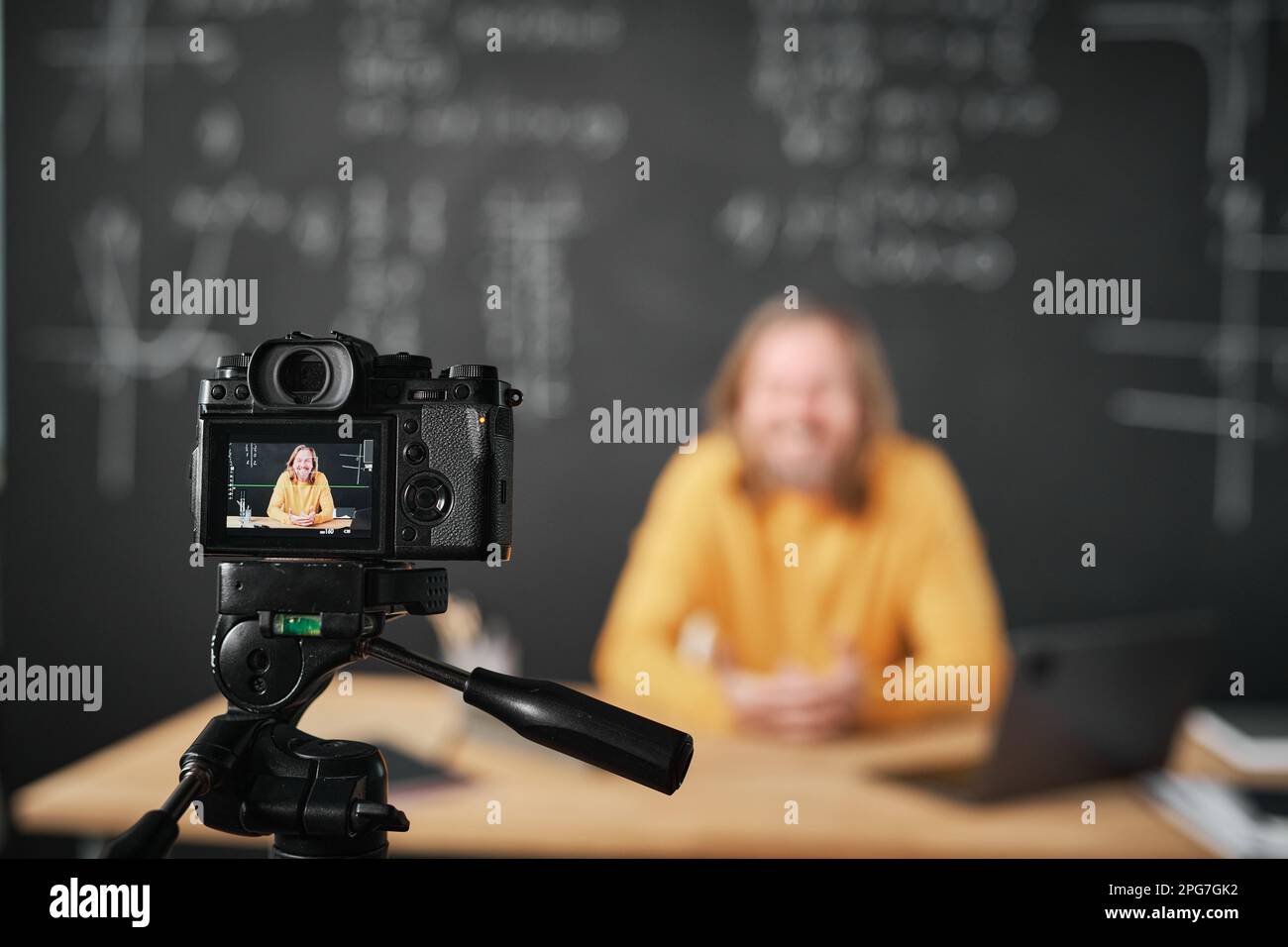 Teacher sitting at table against blackboard and recording lesson on ...