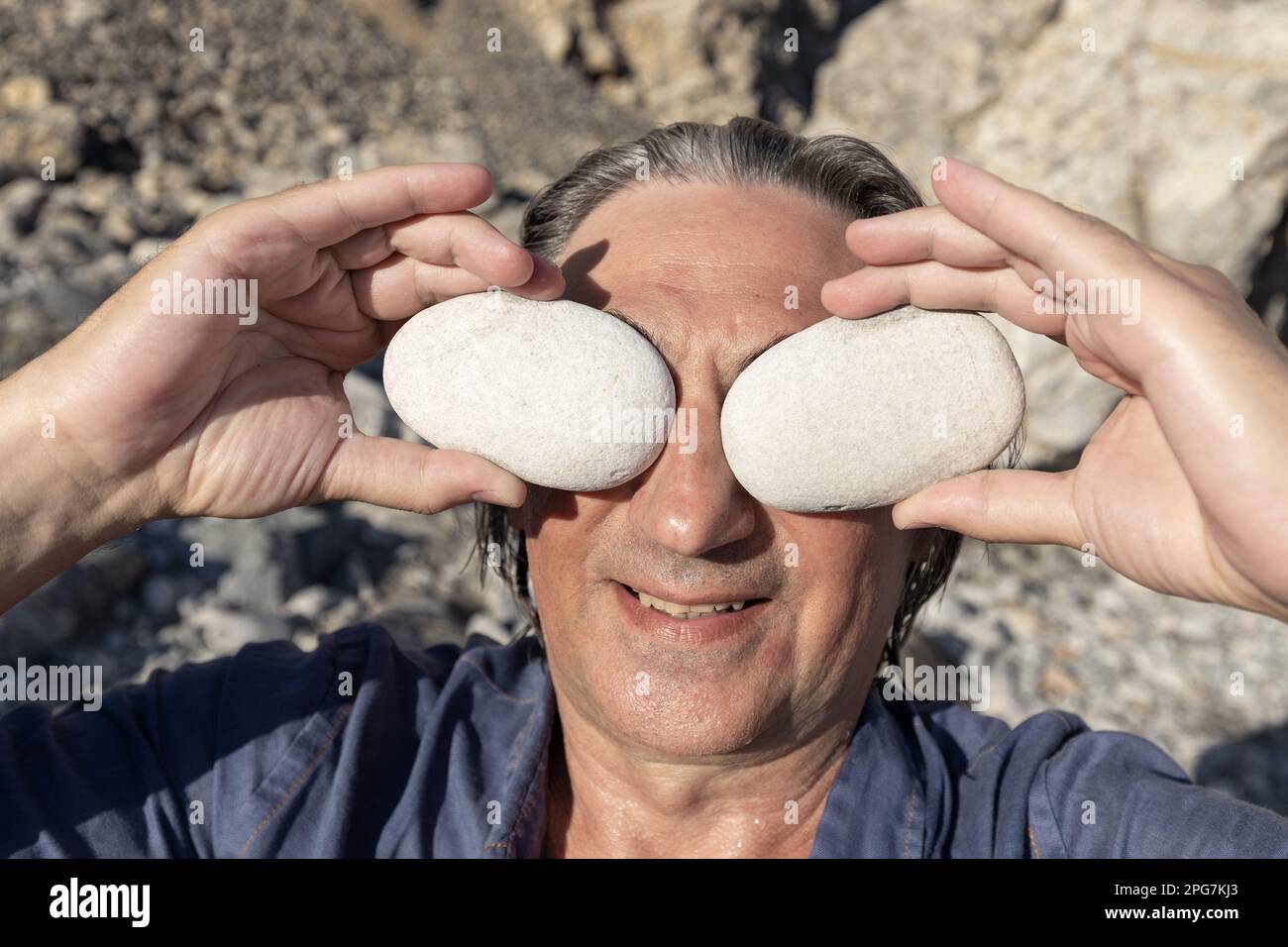A grizzled man on a pebble beach holds oval stones like glasses near ...