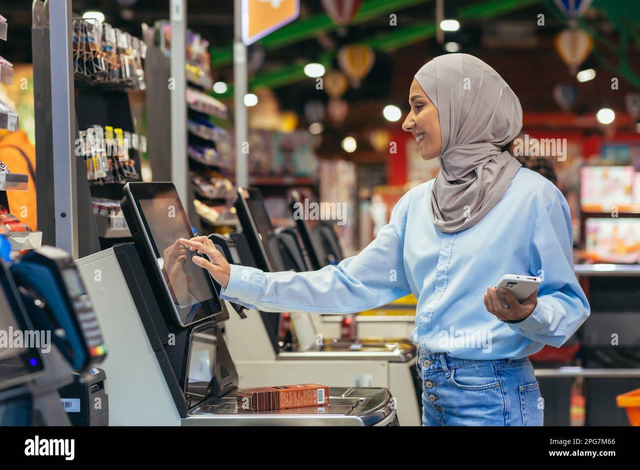A woman buyer in a supermarket in a hijab pays for goods at a self ...