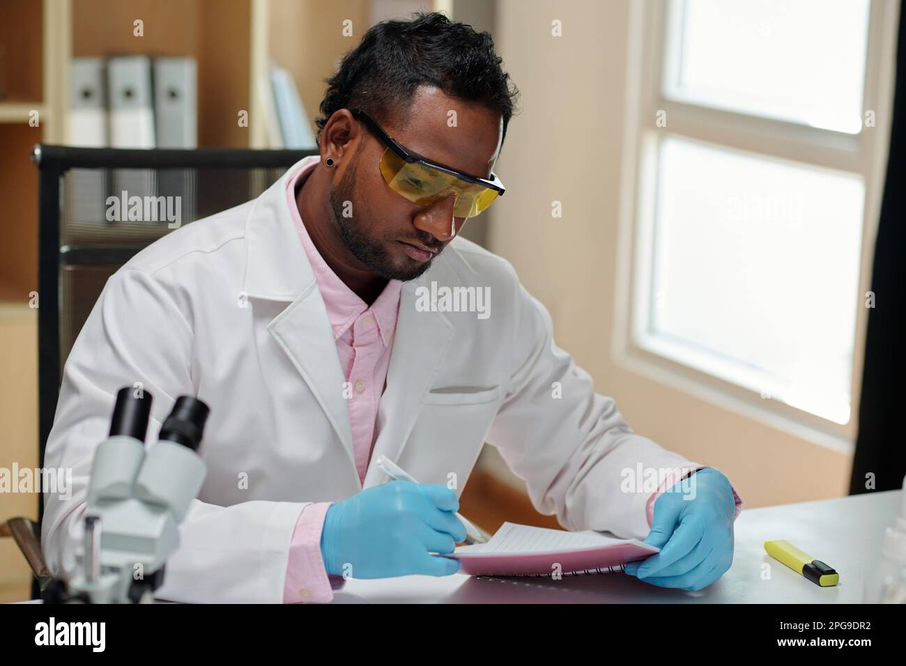 Young man in lab coat and gloves making notes in copybook while sitting ...
