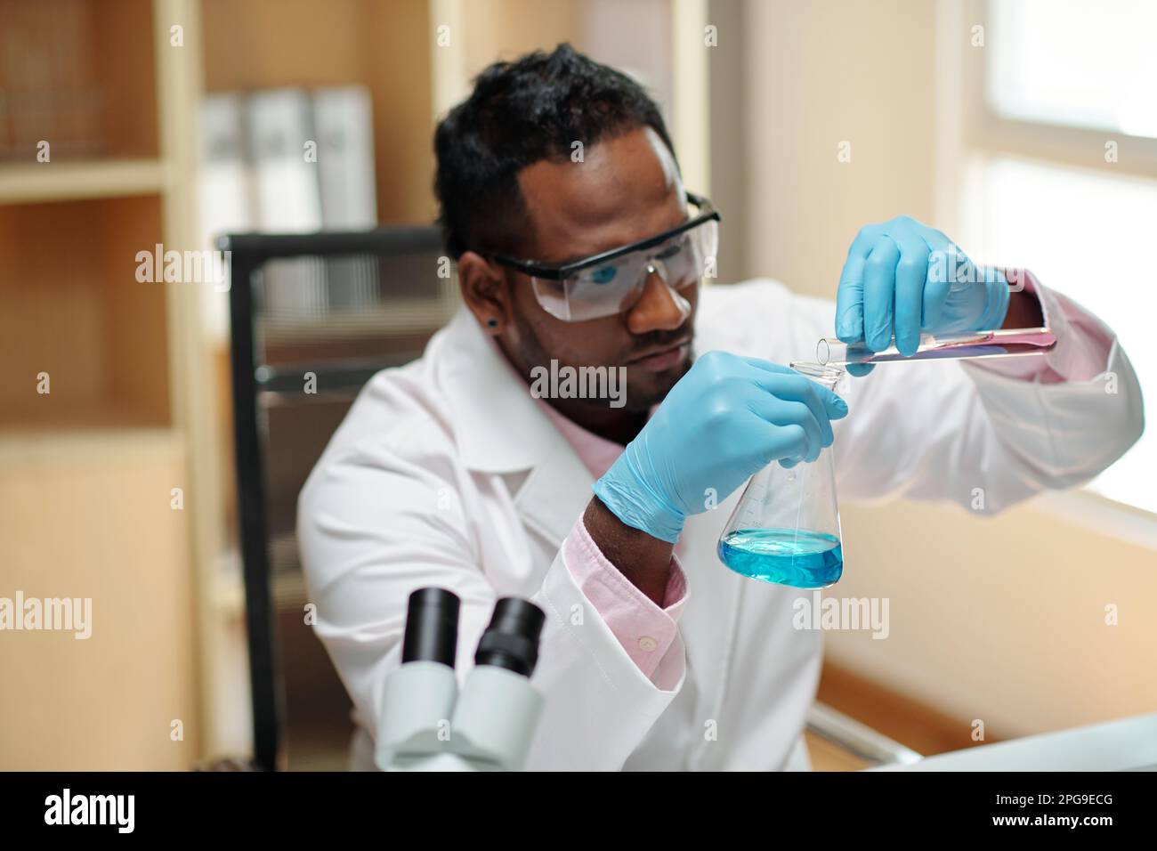 Young black man in workwear pouring liquid from flask into beaker while ...