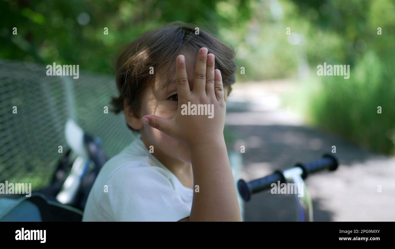 Little boy rubbing nose with hand seated at park bench. Kid touching ...