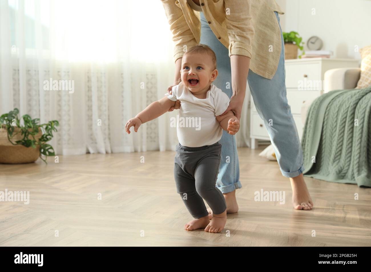 Mother supporting her baby daughter while she learning to walk at home ...