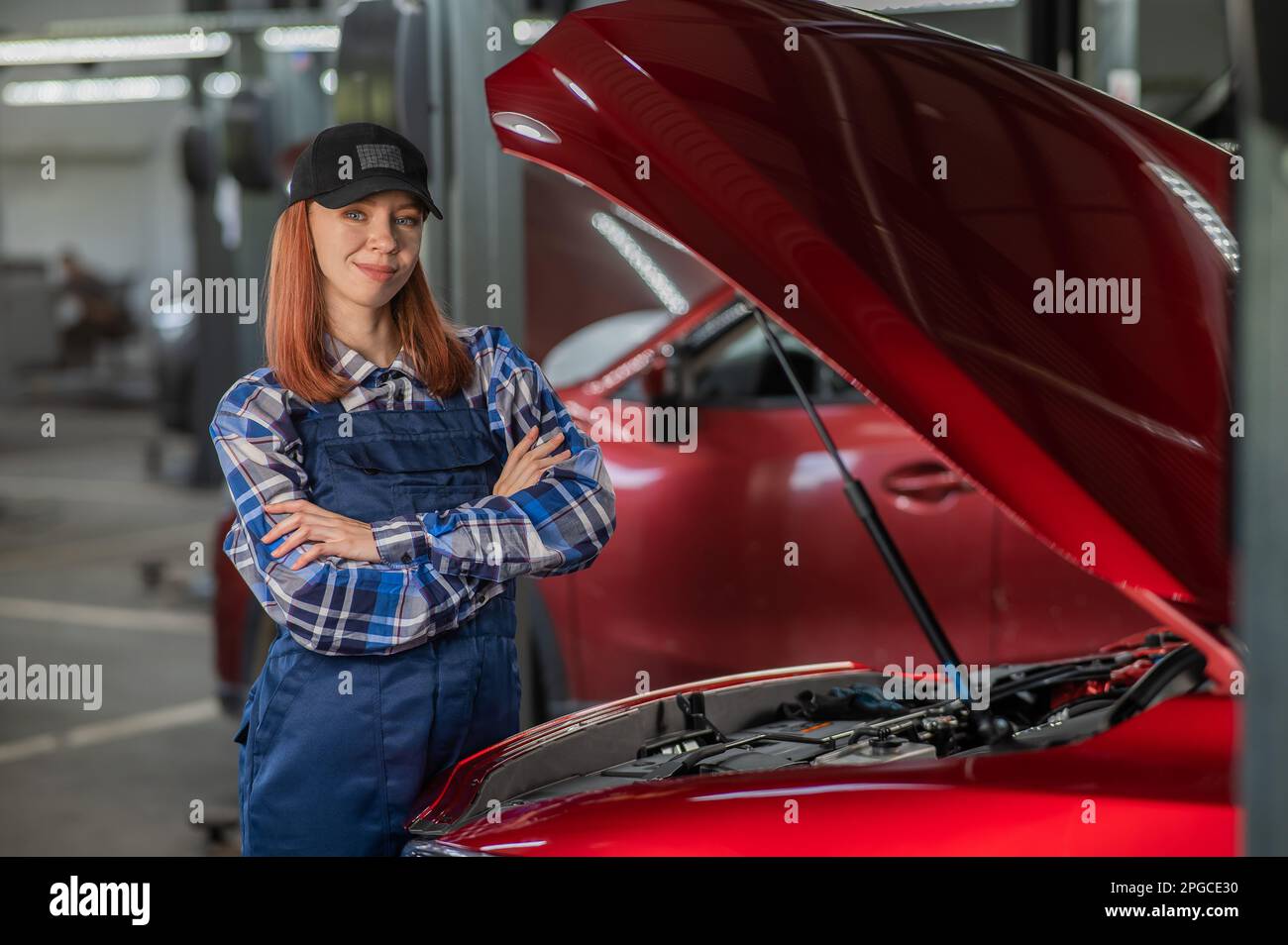 A woman auto mechanic stands at the open hood of a car with her arms ...