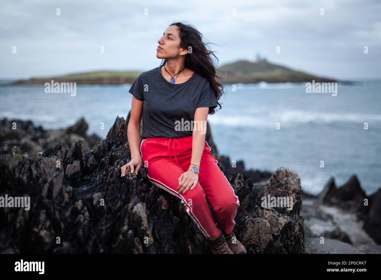 Positive young female sitting on rocky cliff and looking away near wavy ...
