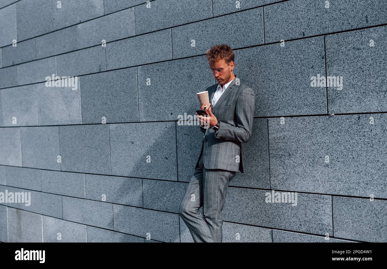 Leans on the wall. Young successful businessman in grey formal wear is ...