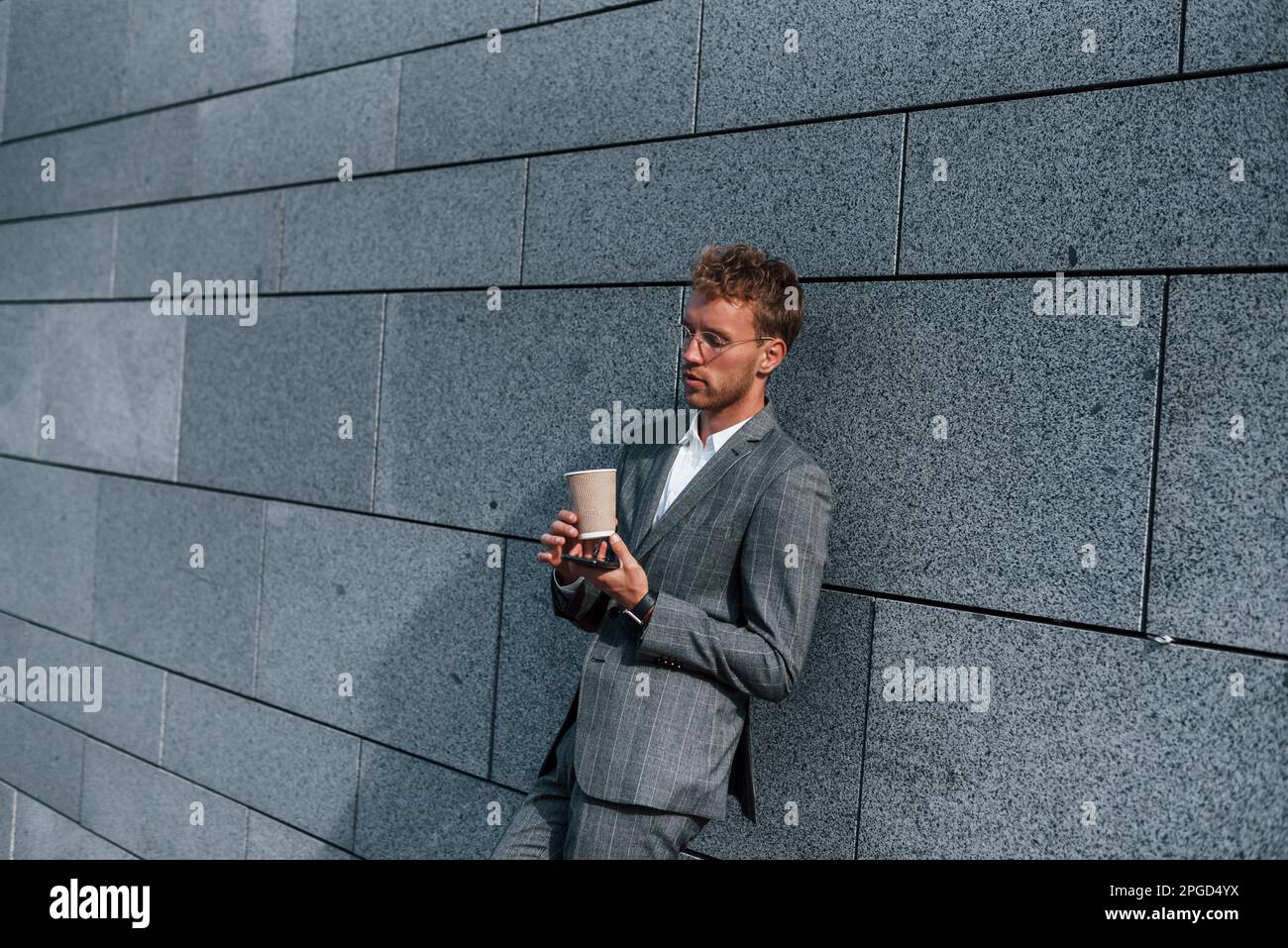 Leans on the wall. Young successful businessman in grey formal wear is ...