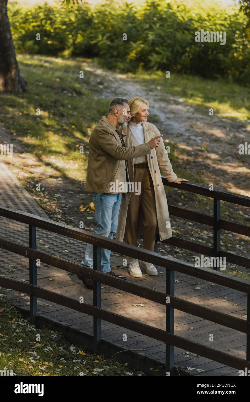 Smiling mature man pointing with finger near blonde wife while standing ...