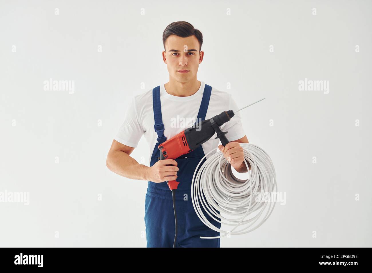 With drill and cables in hands. Male worker in blue uniform standing ...
