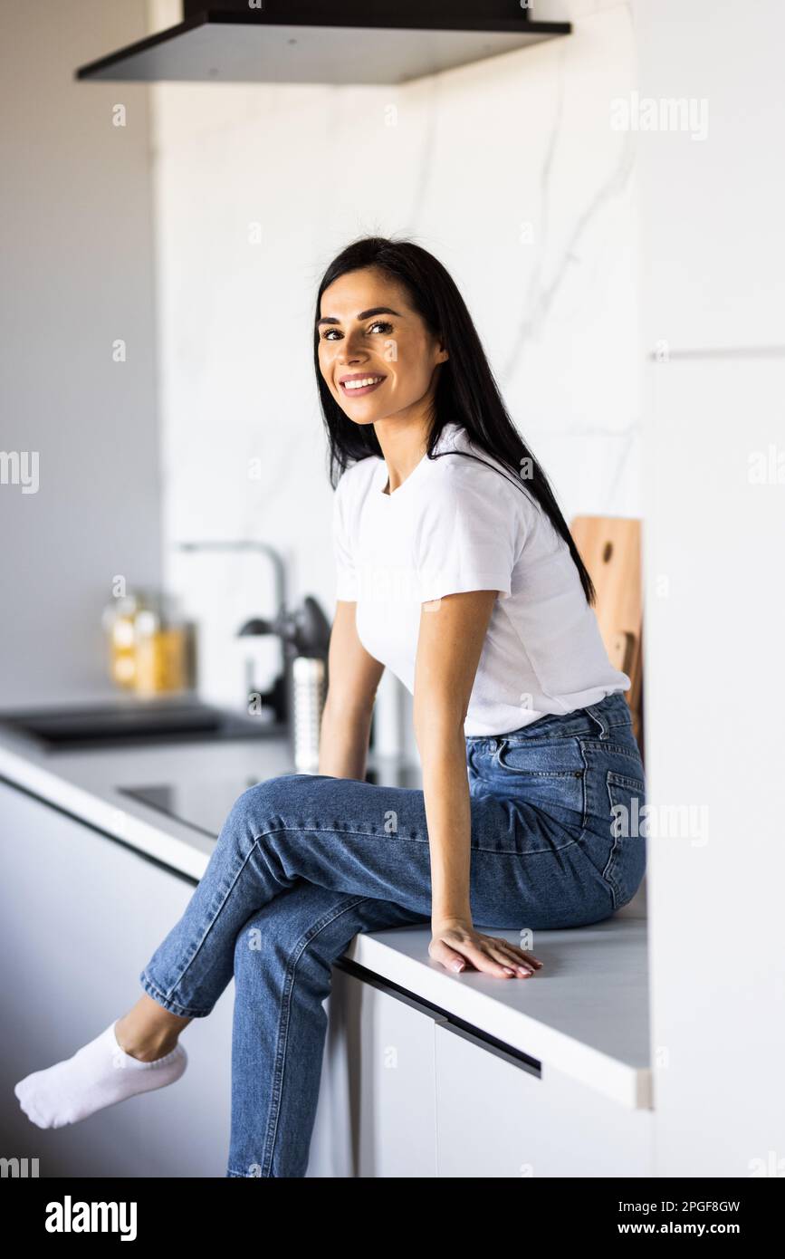 Portrait of beautiful woman relaxing sitting on counter top at kitchen ...