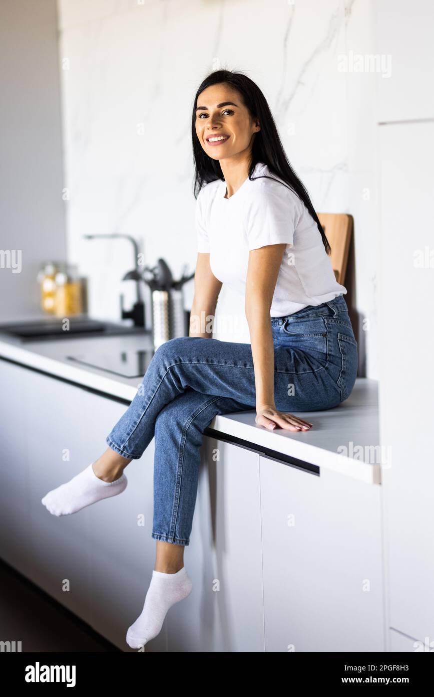 Portrait of beautiful woman relaxing sitting on counter top at kitchen ...