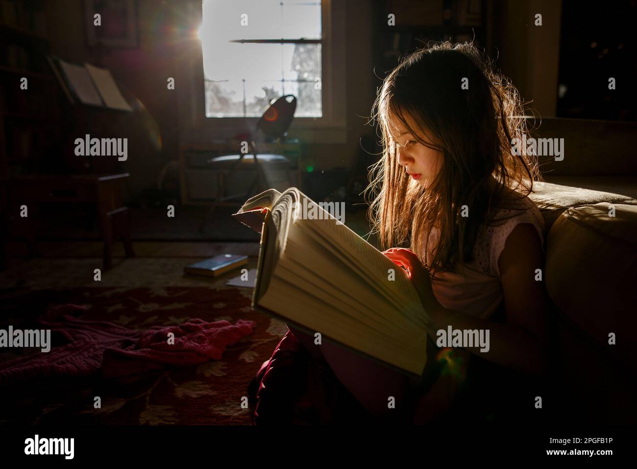A small child sits in patch of bright sunlight reading a large book ...
