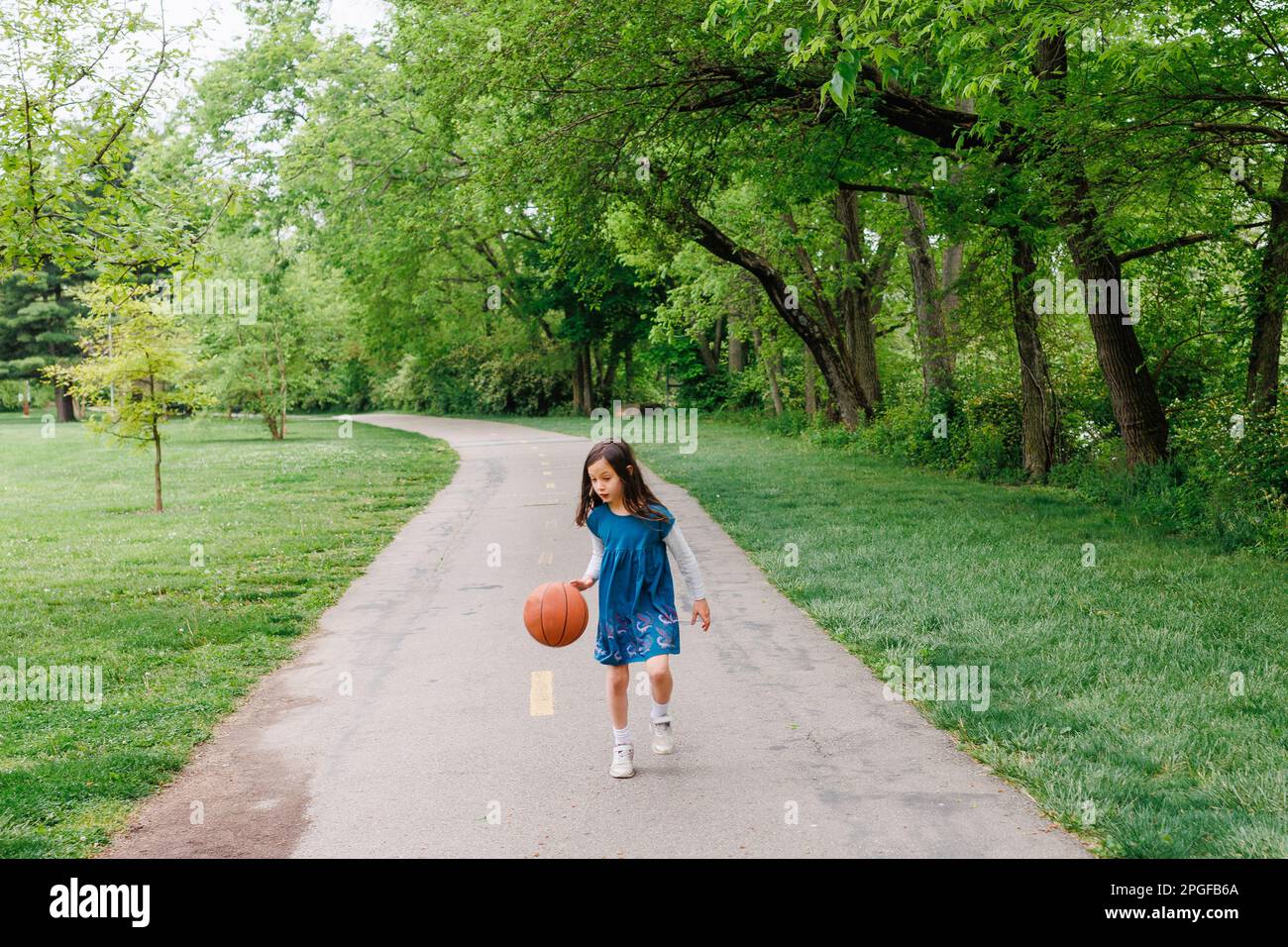 A little girl dribbles basketball alone down long wooded pathway Stock ...