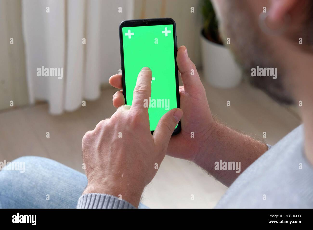 Young man sitting at home holding smartphone green mock-up screen in ...