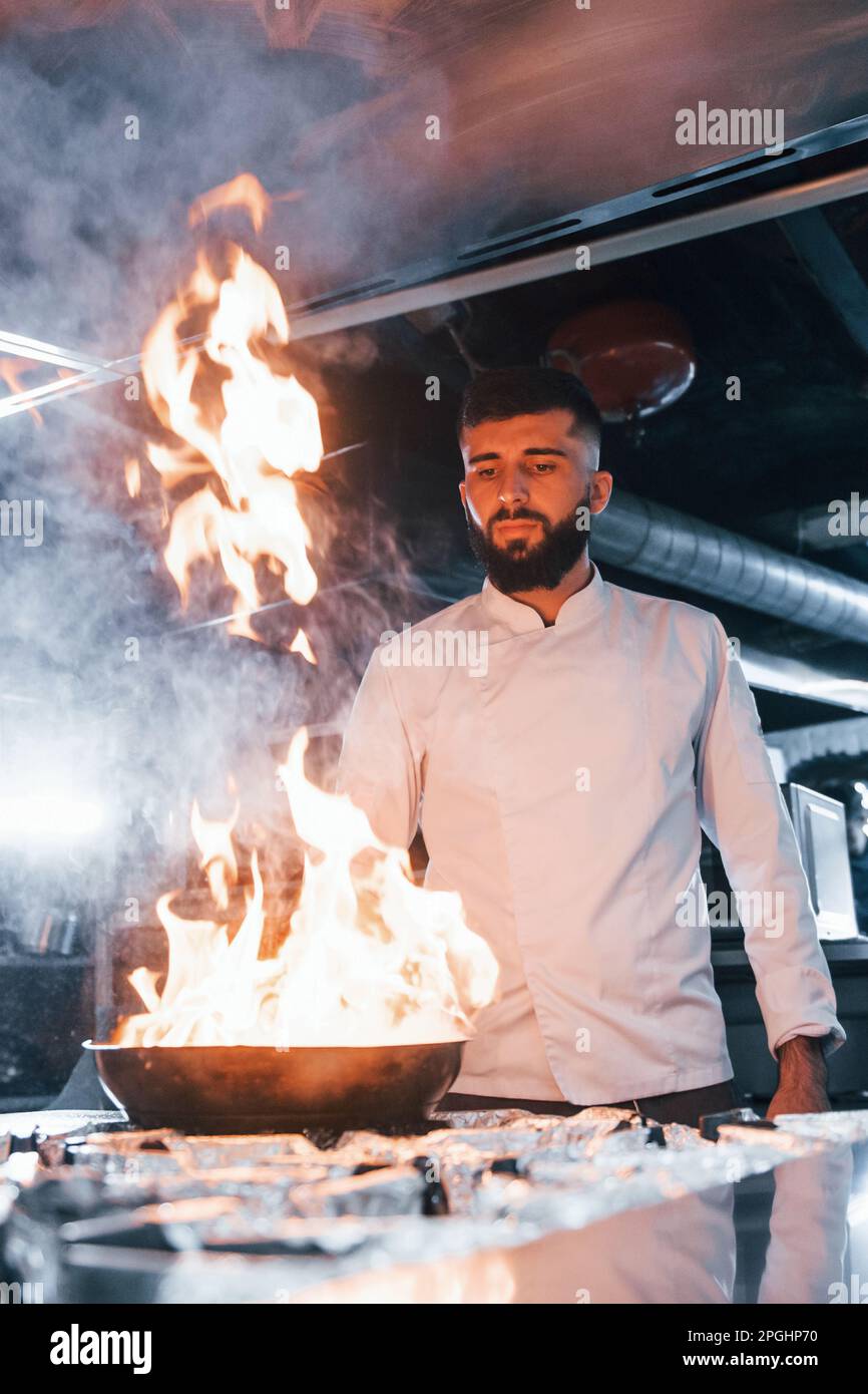 Uses frying pan. Chef in white uniform cooking food at kitchen. Busy ...