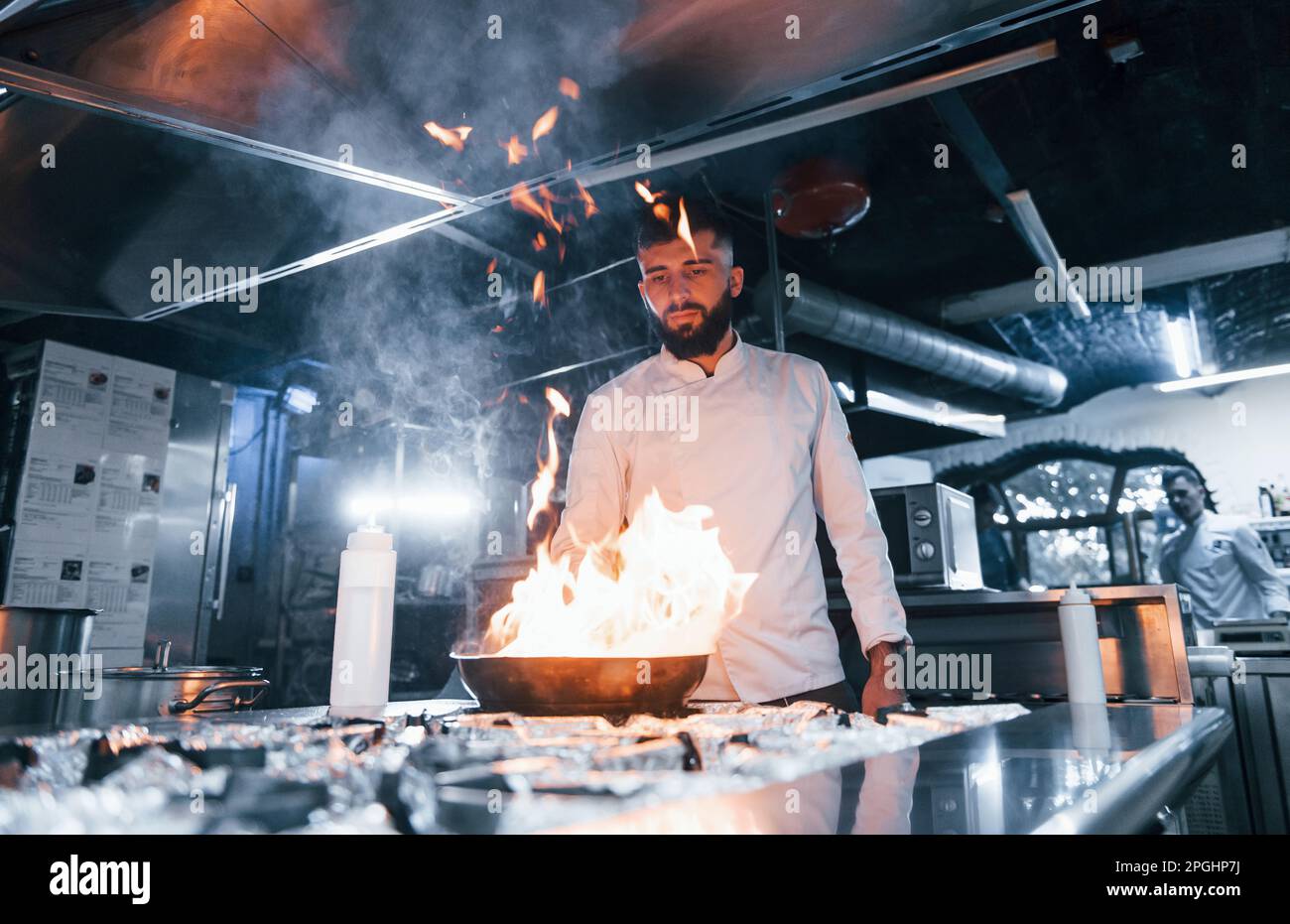 Uses frying pan. Chef in white uniform cooking food at kitchen. Busy ...