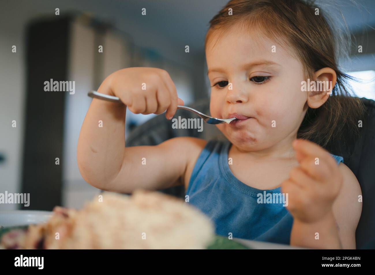 A toddler gir eating with a fork and sitting on a highchair at home ...