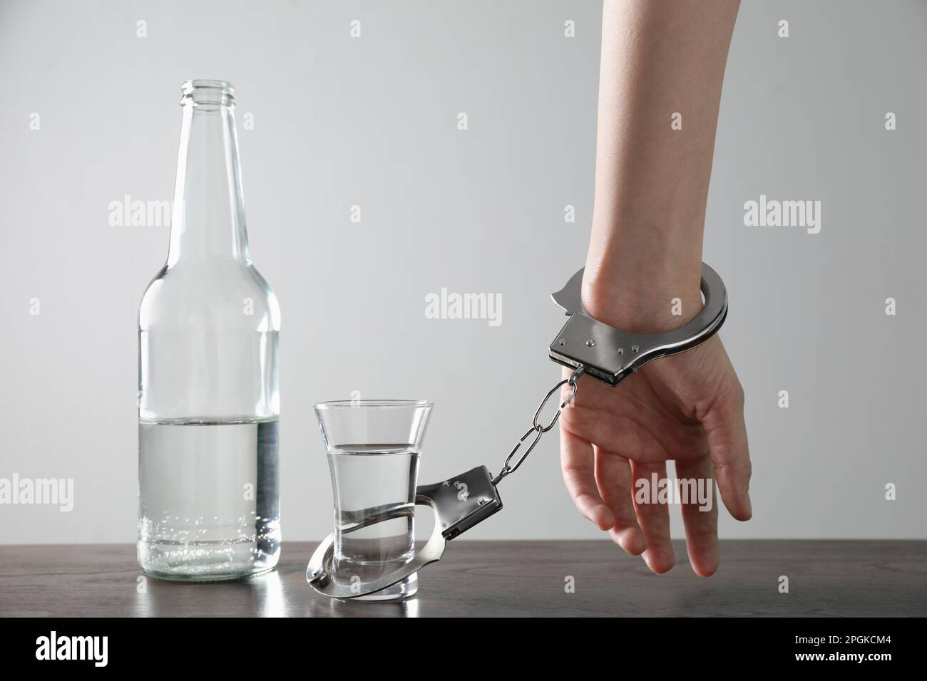 Woman in handcuffs with glass of vodka at wooden table against white ...