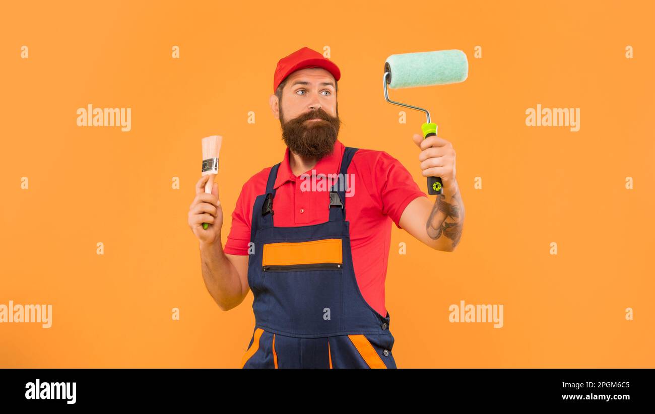 puzzled bearded man in work clothes hold paint roller and brush on ...