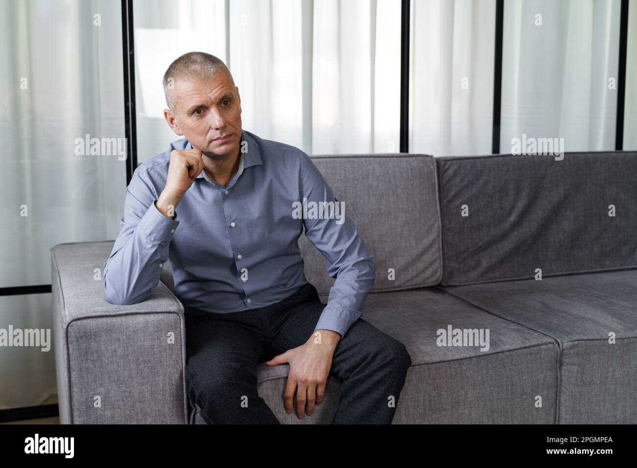 Thoughtful serious man sits on the couch at home, immersed in thoughts ...