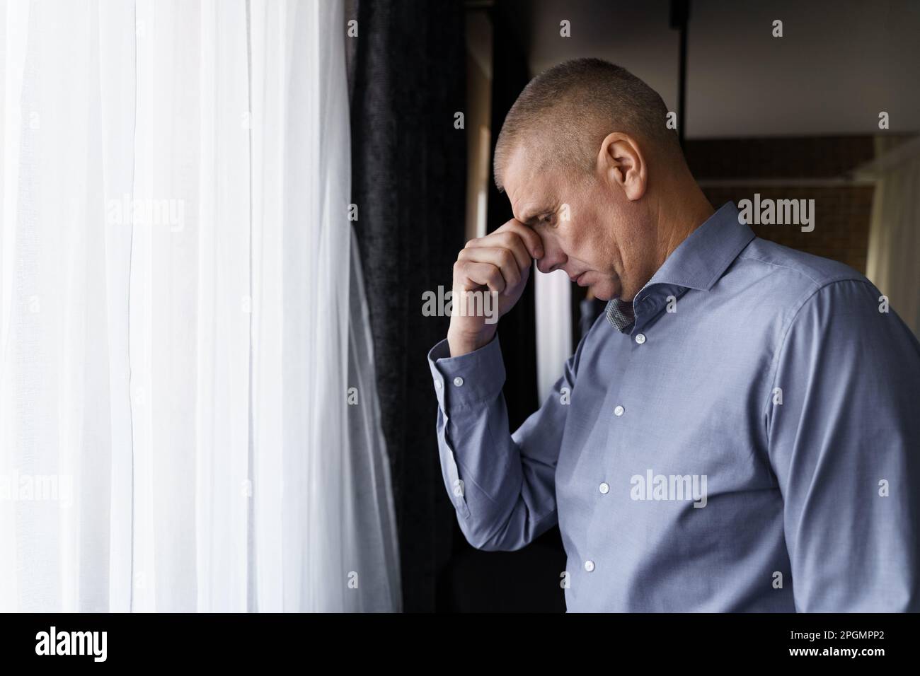 Portrait of a man who stands near the window and holds his head with ...