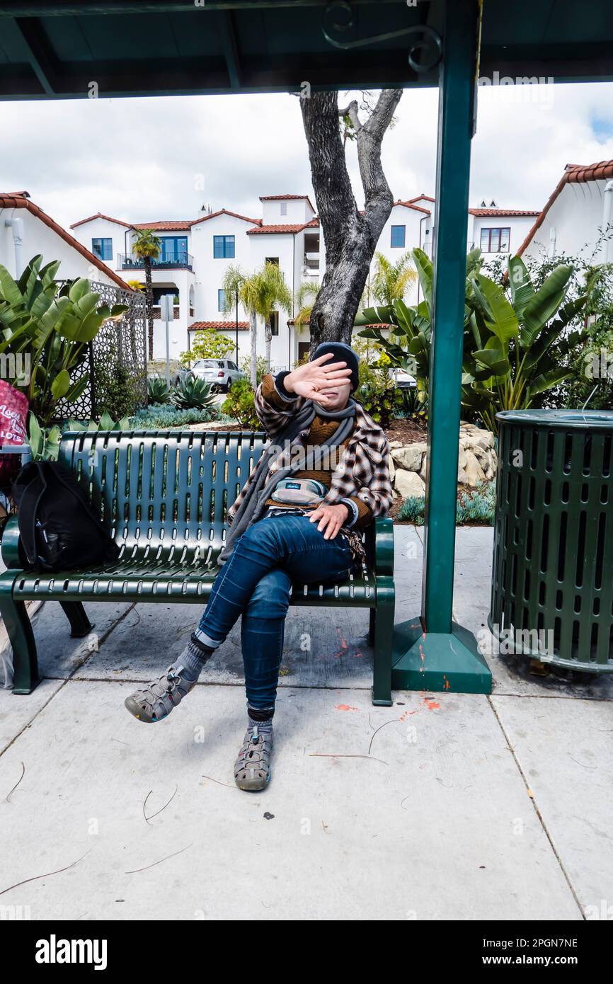 A homeless male sits on a bus stop bench with worldly possessions in a ...