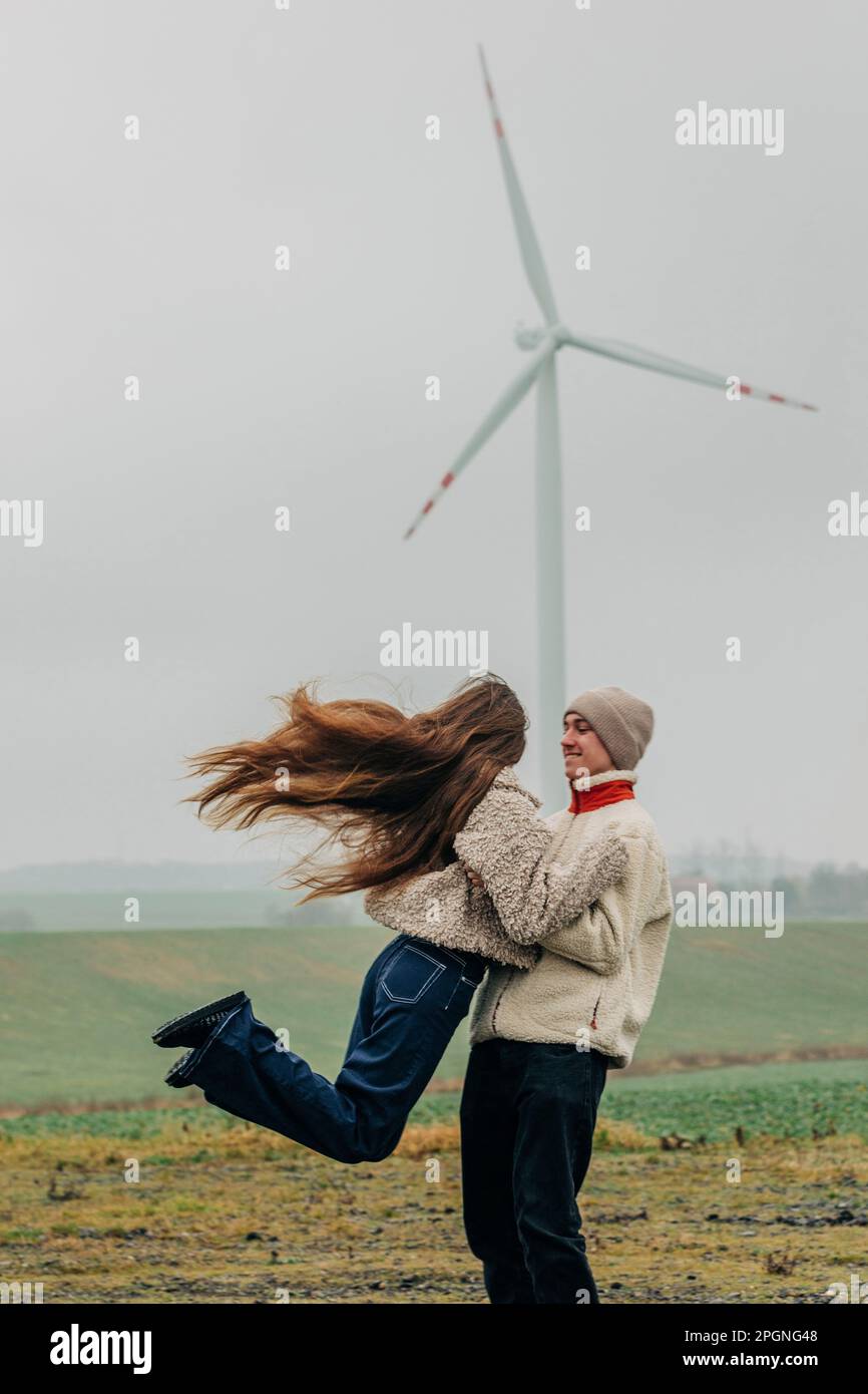 Happy teenage boyfriend spinning girlfriend in front of wind turbine ...