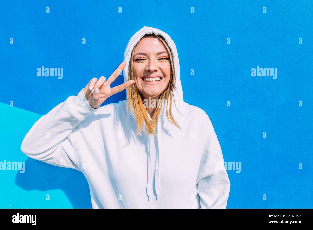 Happy woman making peace sign in front of blue wall Stock Photo - Alamy